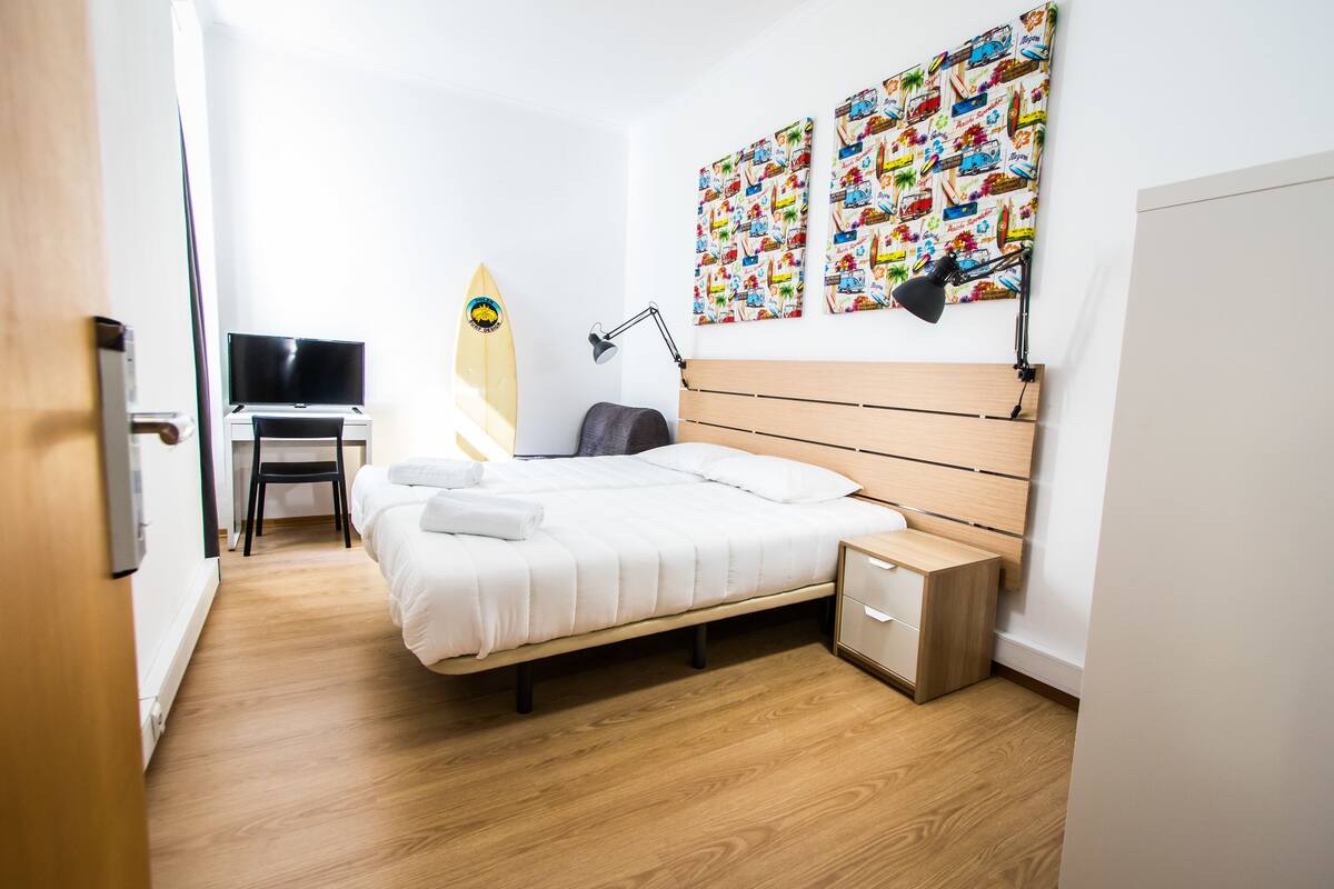 Quarto bem arrumado com cama, mesa de cabeceira, luminária e uma prancha de surf na parede.