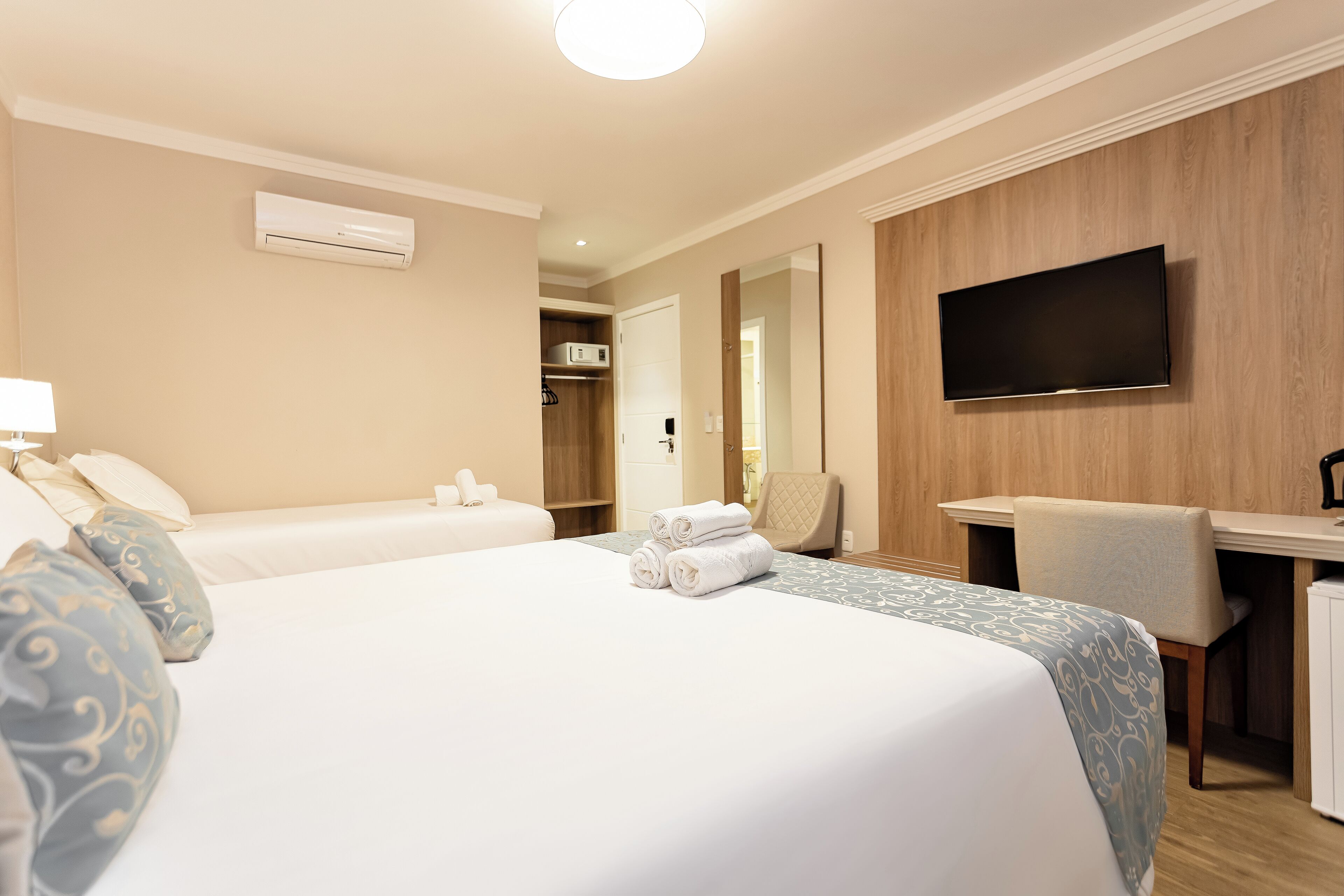 Quarto de hotel com uma cama grande, uma escrivaninha, uma cadeira, uma televisão e um ar condicionado.