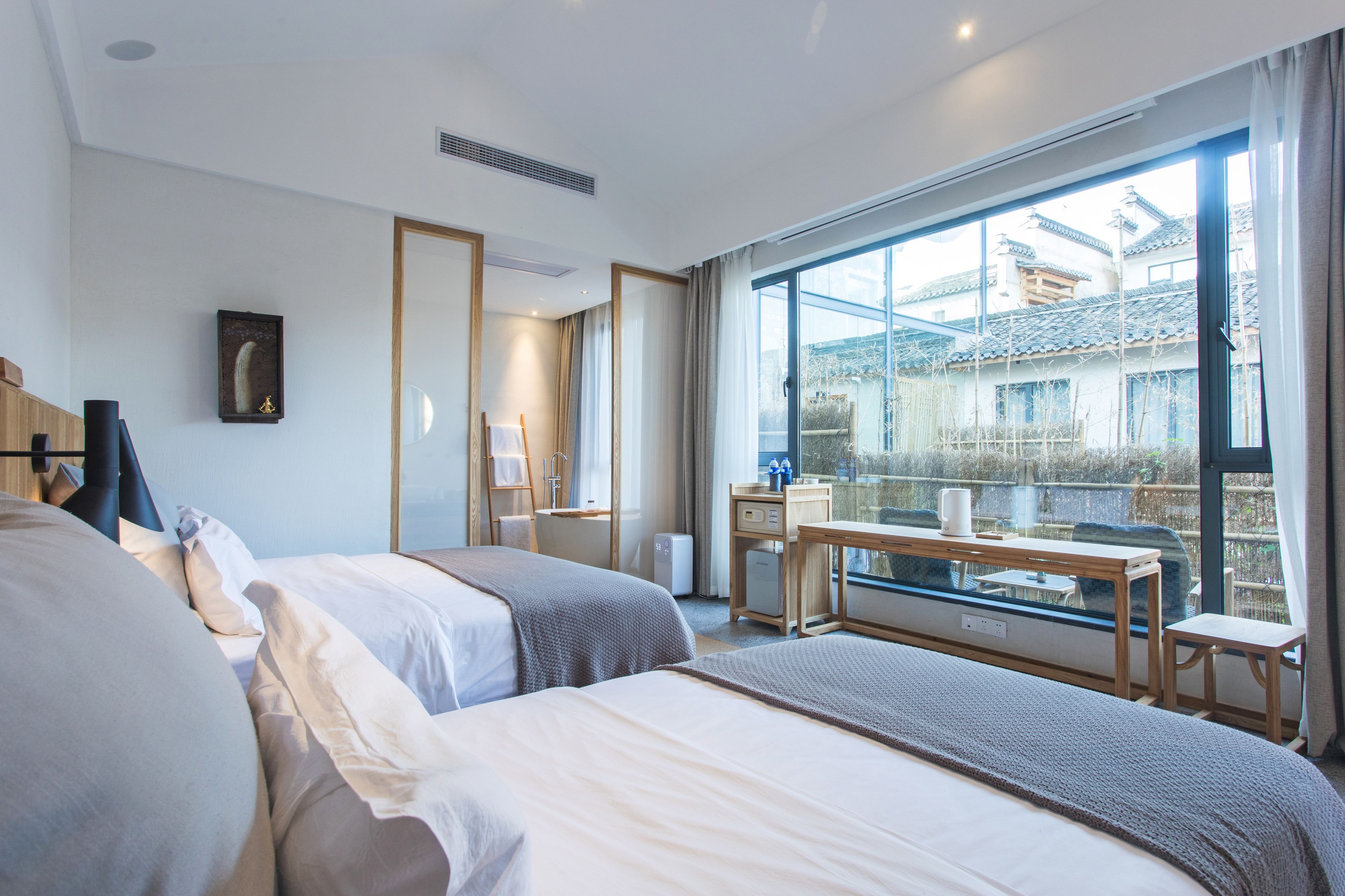 Quarto de hotel com duas camas, uma escrivaninha, uma cadeira e uma janela ampla com vista para arquitetura tradicional.