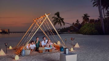 Cena de praia com estrutura de bambu decorada com luzes de fada, palmeiras e um pôr do sol ao fundo.