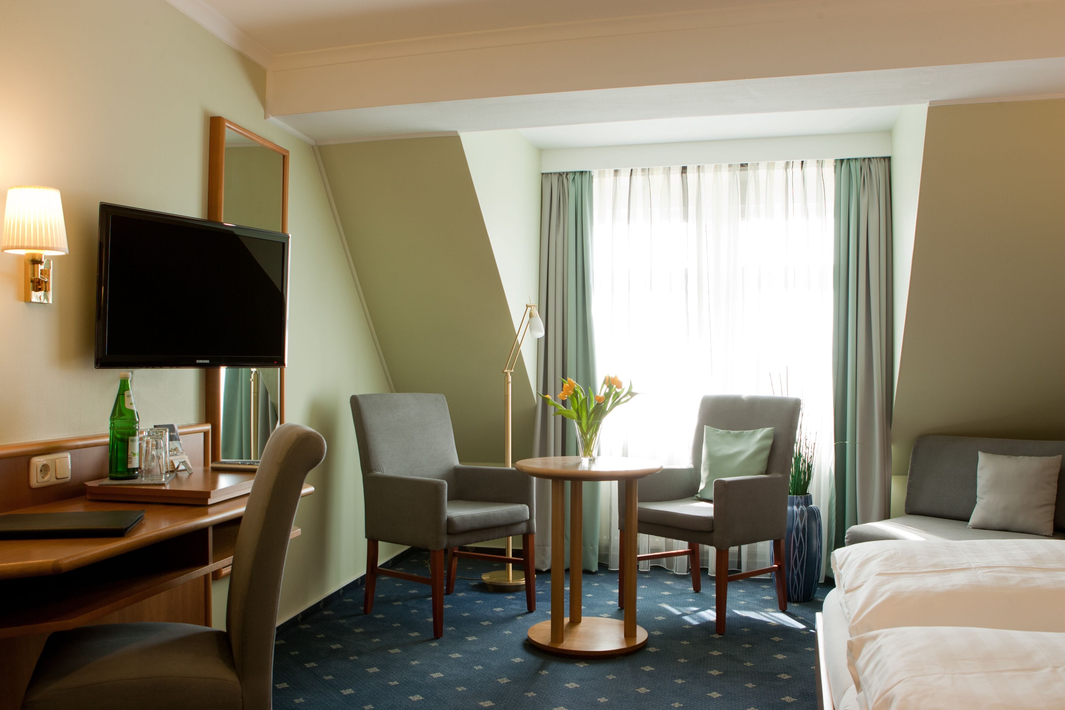 Quarto de hotel com cama, duas poltronas, escrivaninha com TV e pequena mesa com um vaso de flores.