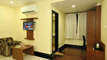 Quarto de hotel com sofá, uma pequena mesa com cesta e garrafas, televisão de tela plana na parede, armário de madeira e porta que dá para outro quarto.
