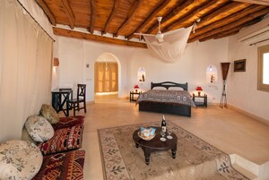 Quarto com teto de madeira, uma cama, mesa de cabeceira com abajur, uma cadeira, sofá e uma pequena mesa com garrafa e copos.