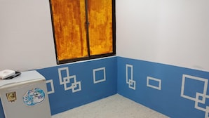 Quarto pequeno com frigorífico, paredes azuis com padrões geométricos brancos, janela com portadas de madeira e chão em azulejos.