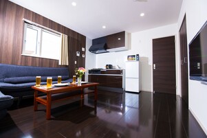 Uma sala de estar moderna com um sofá, uma mesa de centro de madeira com bebidas e uma área de cozinha com armários de madeira escura e uma geladeira branca.