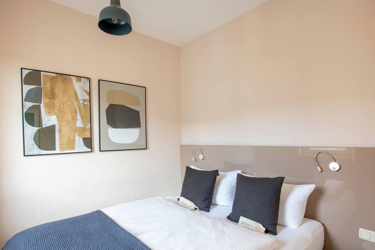 Cama bem arrumada com edredom branco e almofadas escuras, manta azul e arte abstrata na parede.