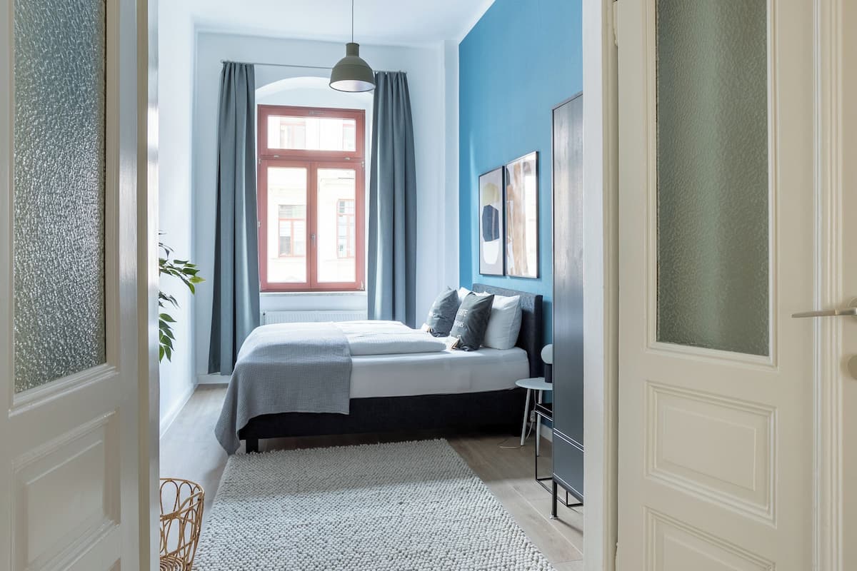 Quarto com cama, janela com cortinas, luminária pendente e porta que dá para outro cômodo.