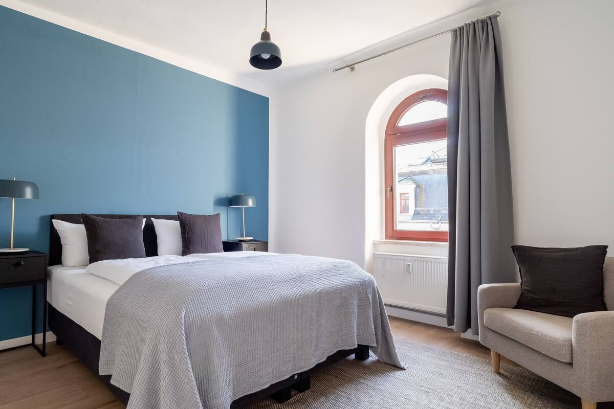 Um quarto moderno com uma cama, mesas de cabeceira, uma cadeira e uma janela com cortinas.