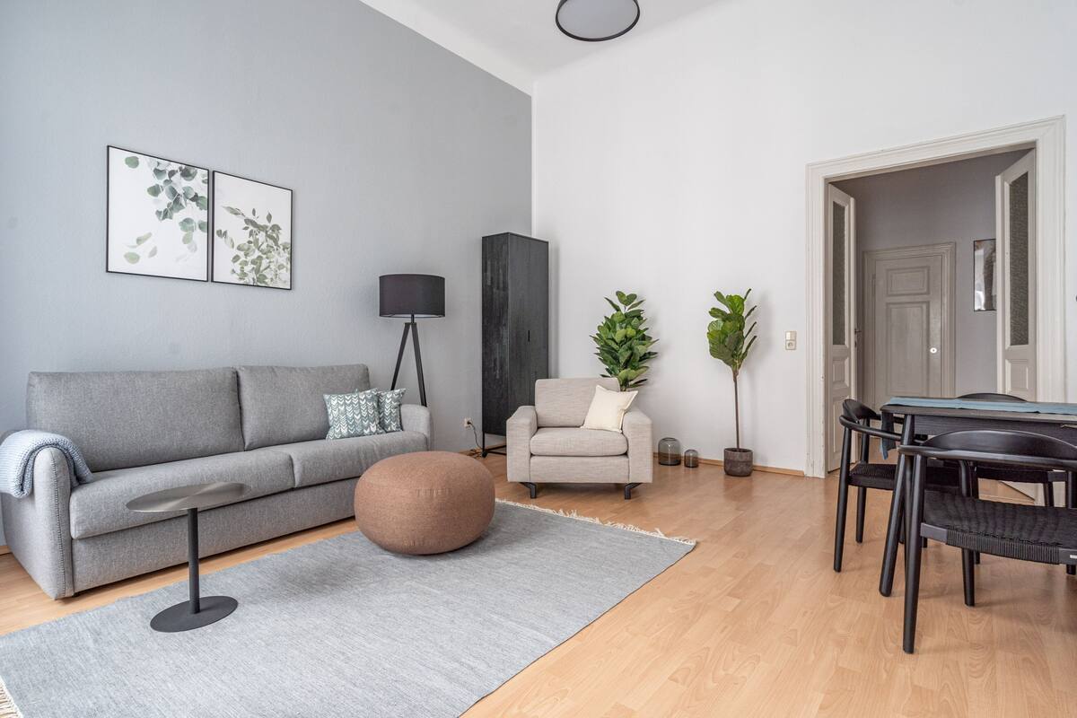 Uma sala de estar moderna com um sofá cinzento, um banquinho redondo, uma mesa de jantar com cadeiras e duas plantas em vasos.