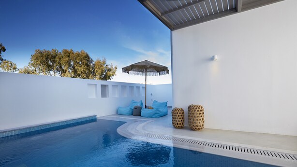 Uma área moderna de piscina ao ar livre com céu azul límpido, um espaço de estar coberto com almofadas e dois bancos decorativos.