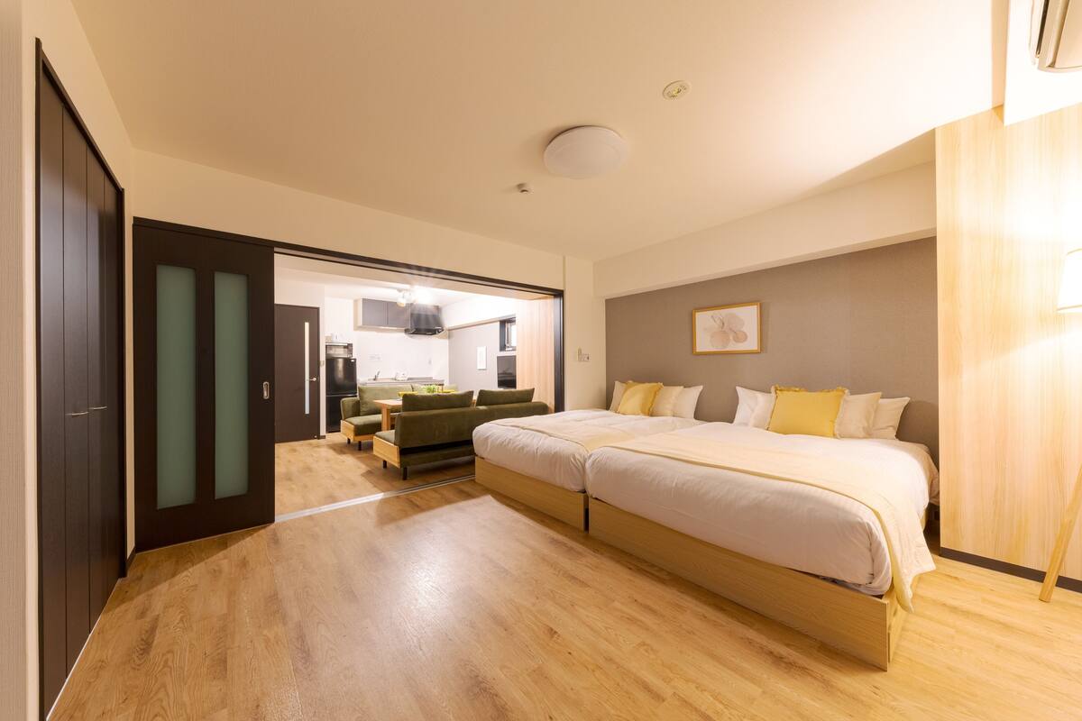 Quarto moderno com cama grande, piso de madeira e cozinha compacta ao fundo.
