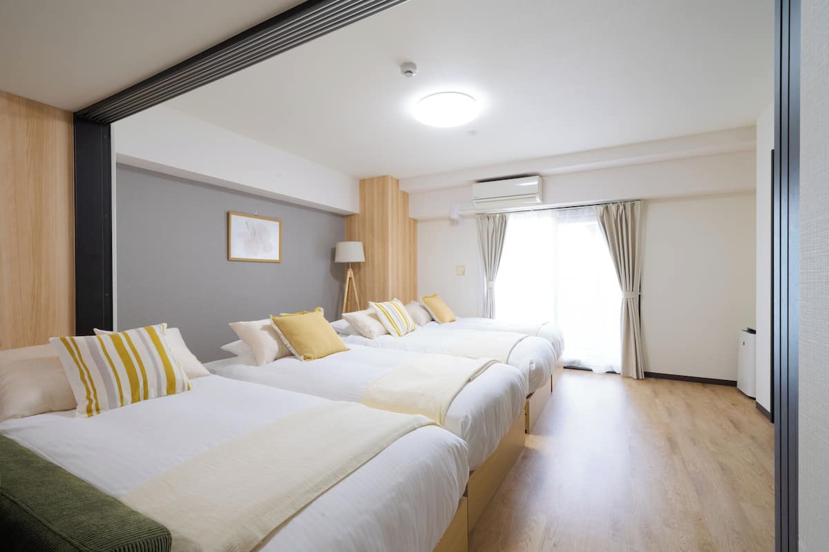 Quarto de hotel com três camas, uma luminária no teto, aparelho de ar condicionado e uma janela com cortinas.
