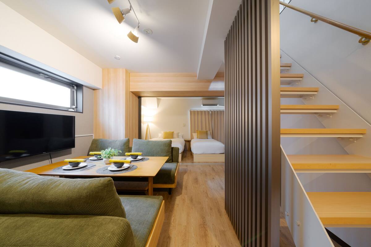Quarto de hotel moderno com piso de madeira, TV de tela plana, área de jantar com mesa e cadeiras, um sofá e uma escada.