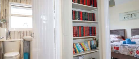 Quarto com um guarda-roupa branco, uma estante cheia de livros, uma cama com um jornal aberto e uma janela com vista para pessoas.