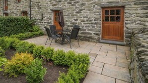 Um edifício de pedra com porta e janelas de madeira, um pátio com mesa e cadeiras, e um jardim com diversas plantas.