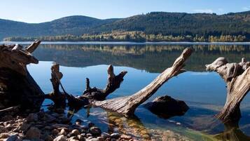 Um lago tranquilo com reflexos nítidos, rodeado por margens rochosas e uma cadeia montanhosa arborizada.