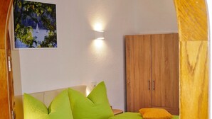 Cama com almofadas verdes e laranja, guarda-roupa de madeira e quadro pendurado na parede.