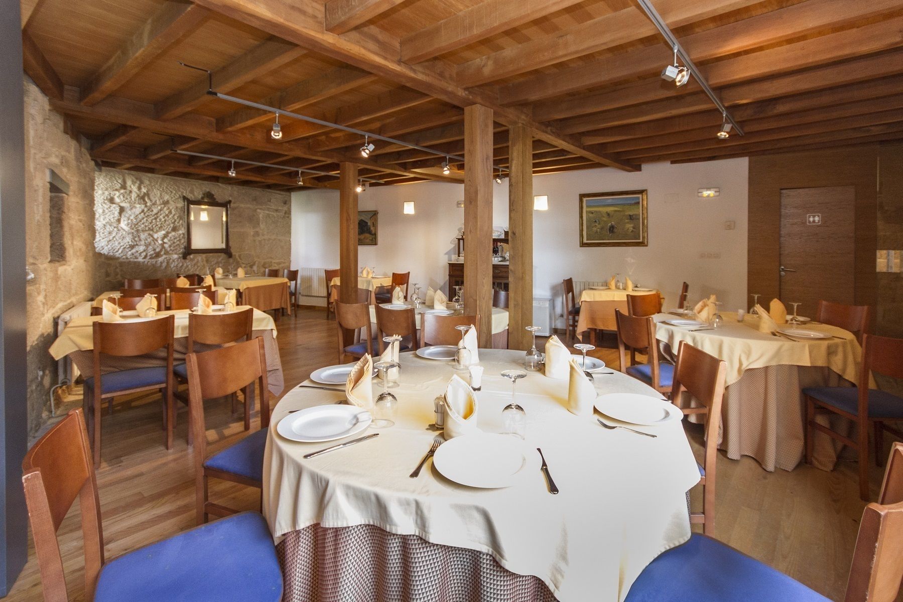 Um restaurante com mesas redondas preparadas para refeições, vigas de madeira no teto, paredes de pedra e uma pintura na parede.