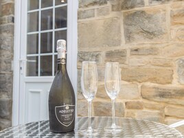 Uma garrafa de vinho Adeleotto Prosecco DOC e dois copos de vinho vazios sobre uma mesa de metal, com um muro de pedra ao fundo.