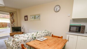 Um quarto aconchegante com uma mesa de madeira e cadeiras, um sofá, um relógio, um quadro emoldurado, uma televisão e um micro-ondas.