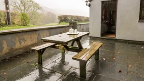 Uma mesa de piquenique de madeira com um banco, um lanterna branca e uma pequena mesa com xícaras e um bule num terraço.