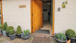Entrada de um edifício com uma porta de madeira, uma placa com a inscrição 'The Milking Parlour' e plantas em vasos na parte externa.