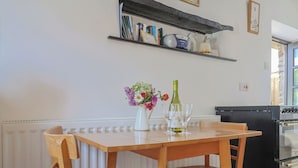 Uma cozinha com uma mesa de madeira, duas cadeiras, um vaso com flores, uma garrafa de vinho e uma estante com utensílios de cozinha.