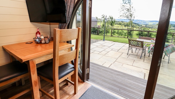 Uma mesa de madeira com uma cadeira, uma televisão na parede e vista para um pátio exterior com mesa e cadeiras.