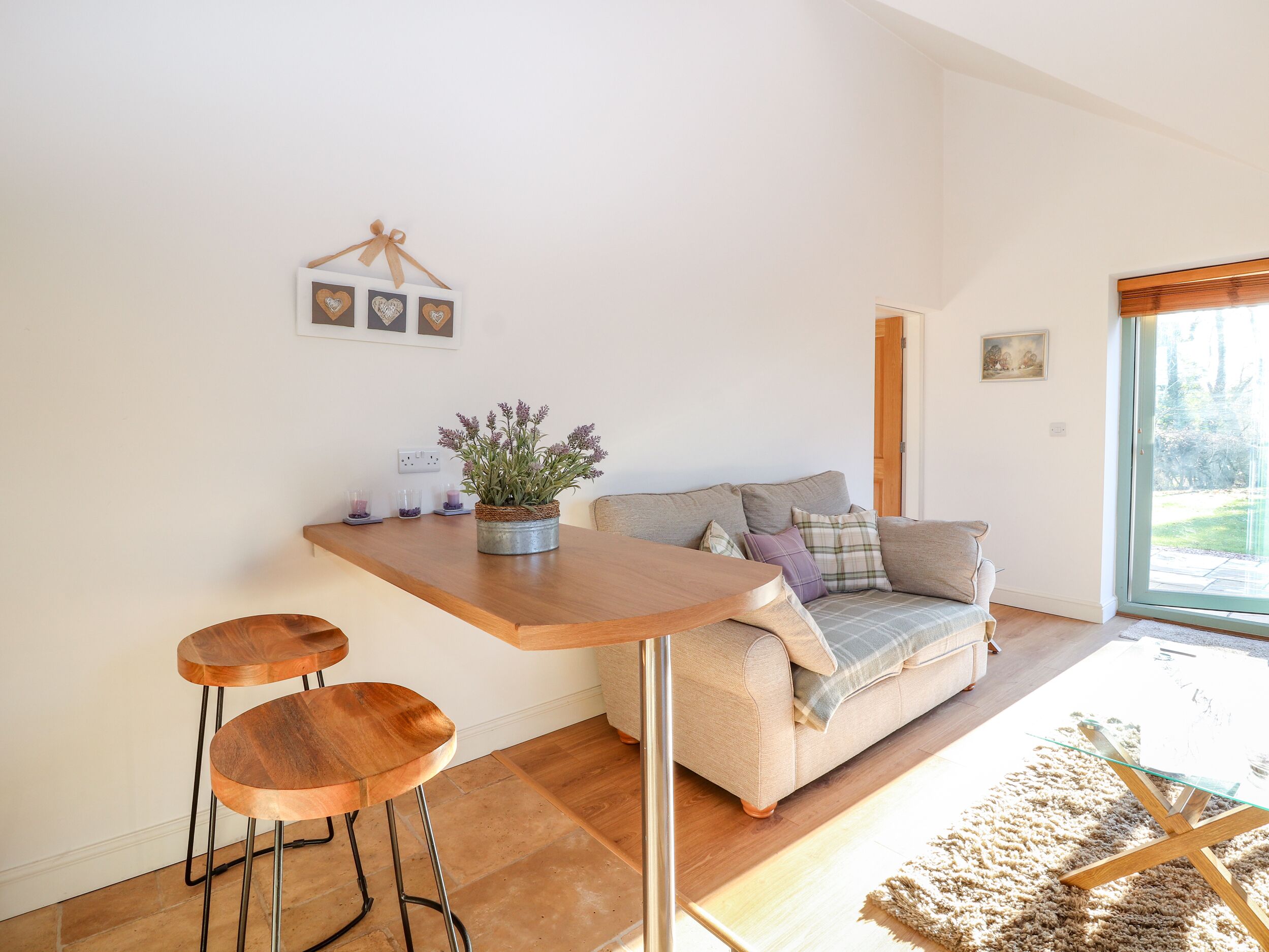 Uma sala de estar moderna com uma mesa de jantar de madeira, dois bancos altos, um sofá bege e uma grande janela com vista para o exterior.
