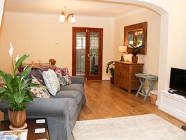 Uma sala de estar com um sofá cinzento, um armário de madeira, um suporte para TV e um espaço de jantar com uma mesa posta para dois.