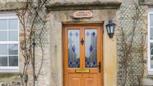 Uma cabana de pedra com porta de madeira, uma placa com o nome "HOPE COTTAGE" e uma janela à esquerda.