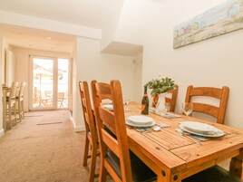 Uma sala de jantar com mesa de madeira posta para quatro pessoas, cadeiras, um vaso com flores e uma garrafa de vinho.