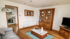Uma sala de estar com um sofá cinza, um armário de madeira com louças, um suporte para TV e uma cozinha visível ao fundo.