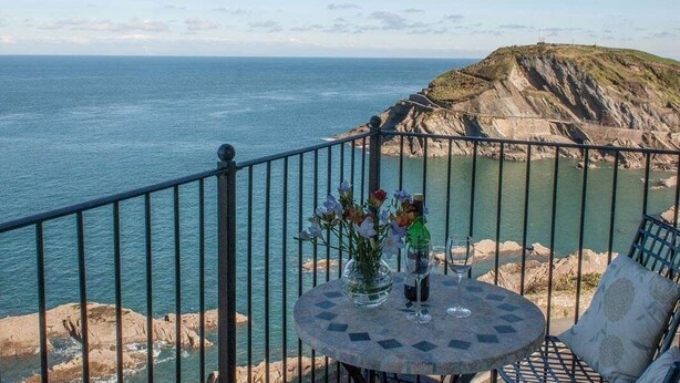Varanda com mesa e cadeiras, vista para a costa com falésias rochosas e o mar.