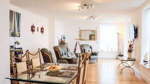 Uma sala de estar ampla com uma mesa de jantar de vidro, poltronas de couro, um espelho grande e uma televisão na parede.