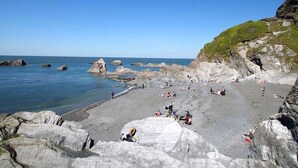 Uma praia rochosa com águas azuis cristalinas, pessoas bronzeando-se e grandes pedras brancas em primeiro plano.