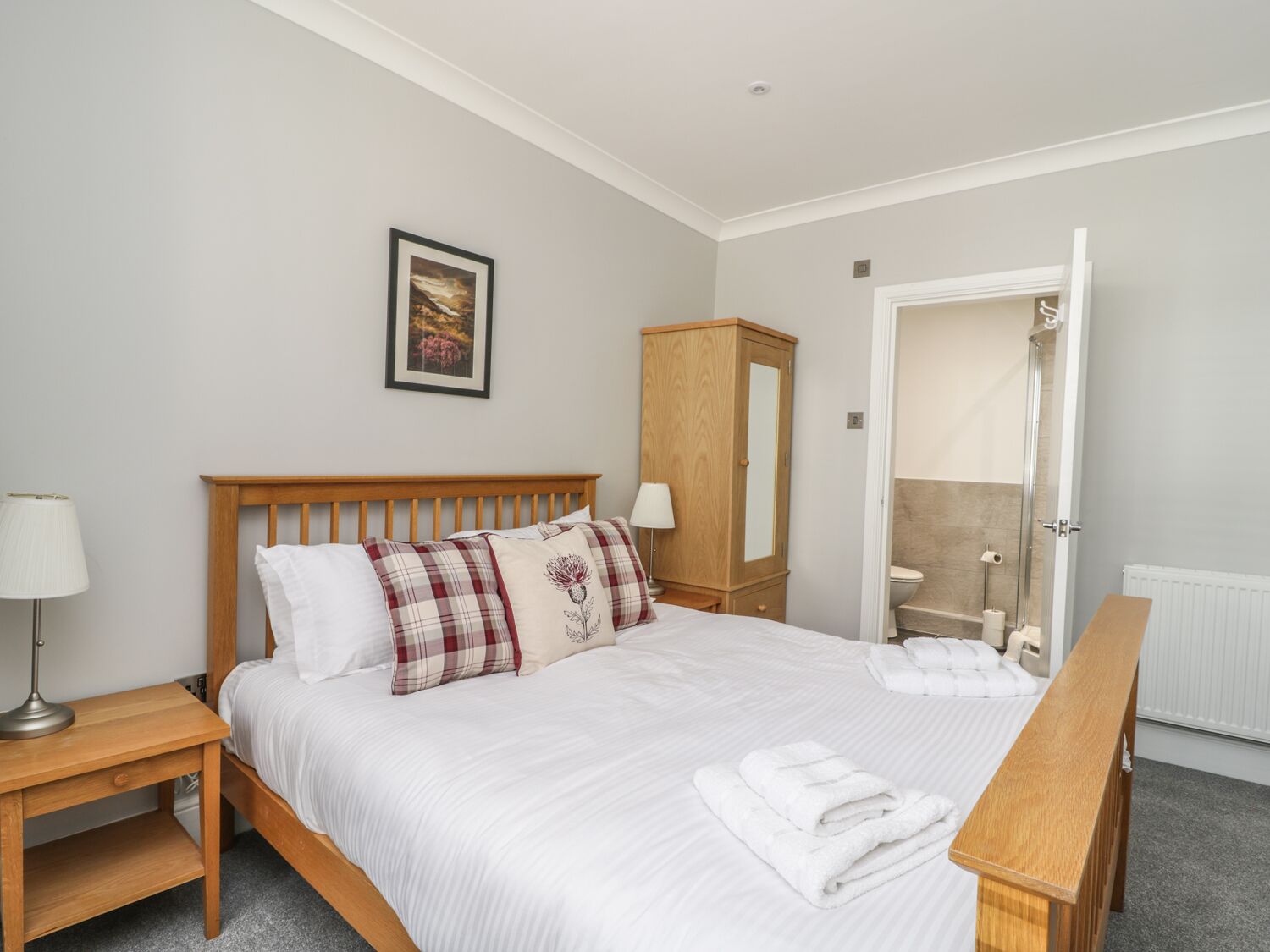 Quarto bem organizado com uma cama, mesa de cabeceira de madeira, guarda-roupa de madeira, banheiro visível através de uma porta aberta e um quadro na parede.