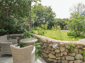 Área de pátio com móveis de vime, parede de pedra e vista para um jardim com diversas plantas e árvores.