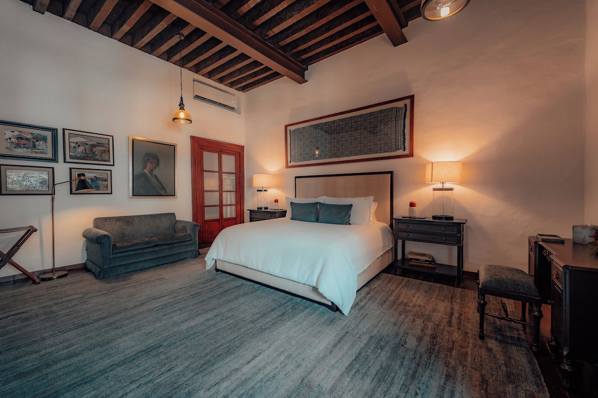 Quarto com cama, sofá, escrivaninha e cadeira. Há luminárias na parede e quadros emoldurados.
