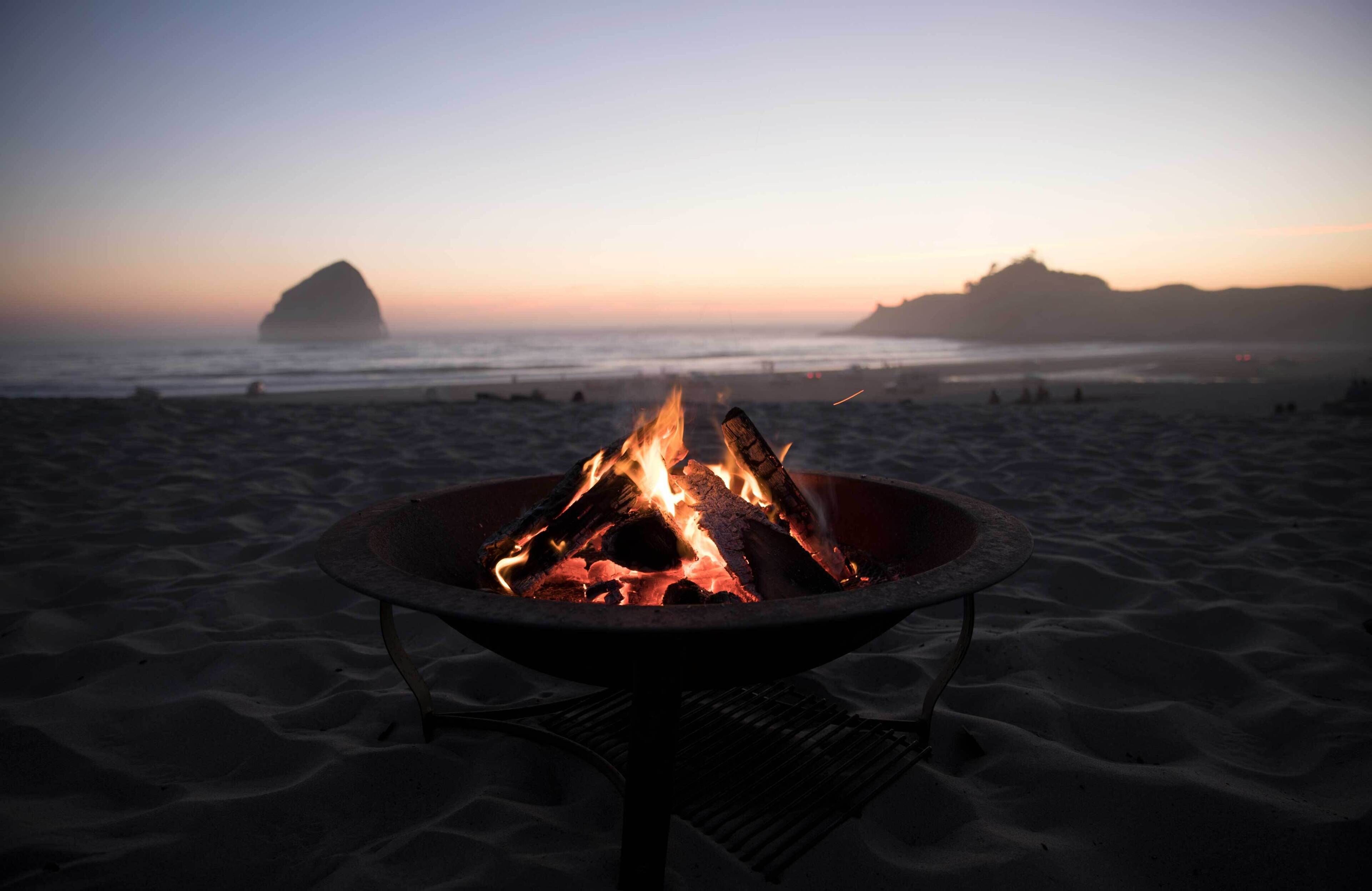 Cena de praia com uma fogueira em primeiro plano e uma grande formação rochosa ao fundo.