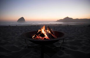 Cena de praia com uma fogueira em primeiro plano e uma grande formação rochosa ao fundo.