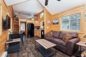 Interior acolhedor de uma cabana, com área de jantar de madeira, sofá e cozinha equipada com frigorífico.