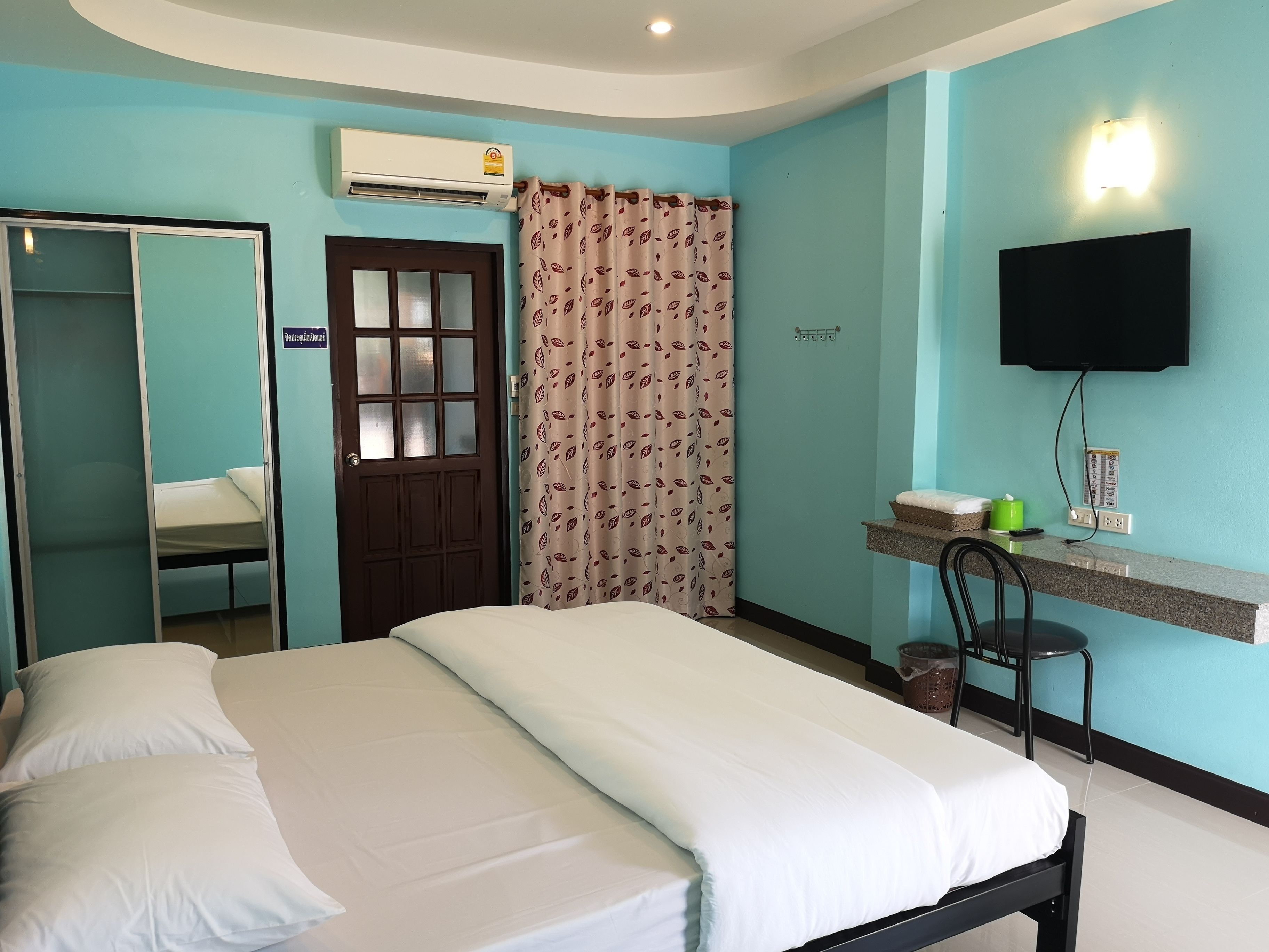 Quarto de hotel com cama, guarda-roupa, ar condicionado, televisão e uma pequena secretária com cadeira.