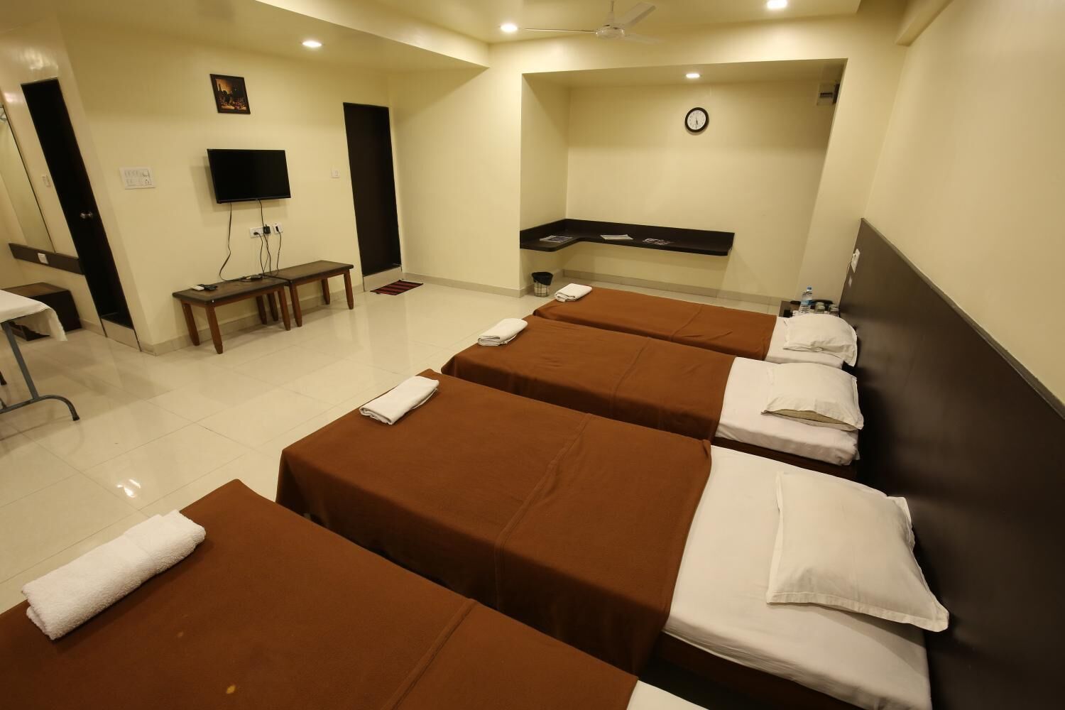 Quarto de hotel com quatro camas, televisão, relógio e pequena mesa de trabalho com cadeira.