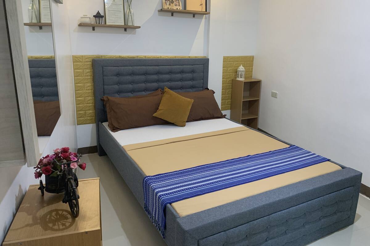 Quarto com uma cama grande, uma mesa de cabeceira, uma prateleira na parede com objetos decorativos e um espelho.