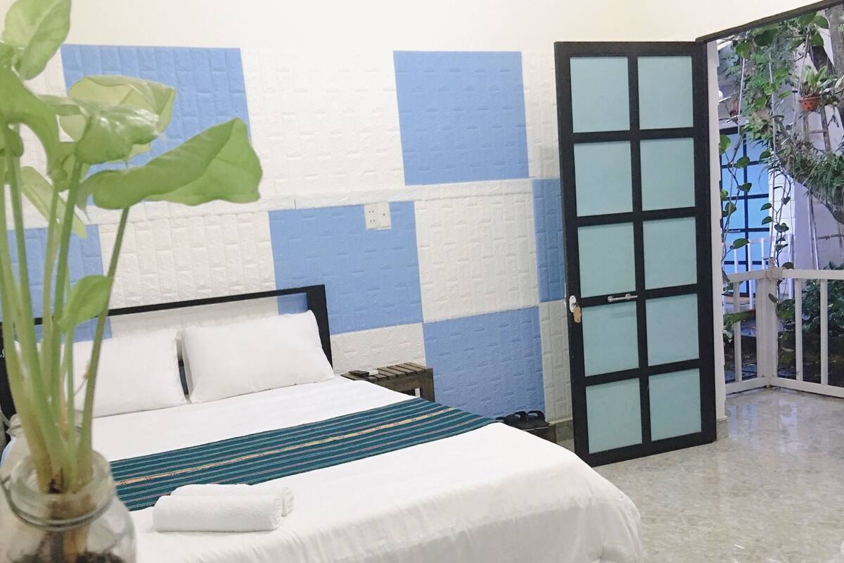 Quarto de hotel com cama, mesa de cabeceira, planta em vaso e vista para a varanda externa.