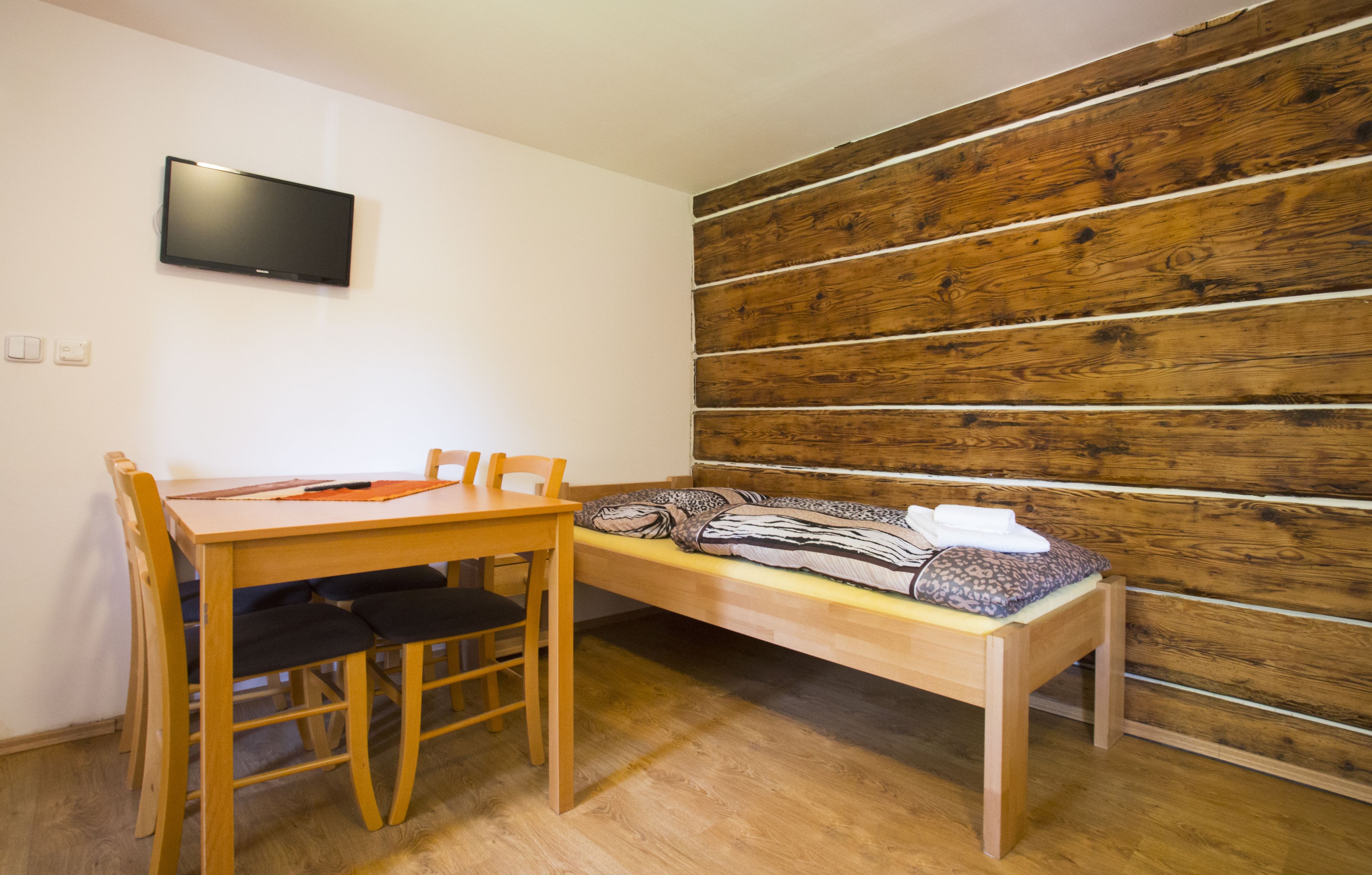 Quarto com cama em madeira estilo banco, mesa de jantar de madeira com cadeiras e televisão de tela plana na parede.