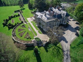 Vista aérea de um grande edifício histórico com um jardim formal, uma entrada com calçada e carros estacionados.