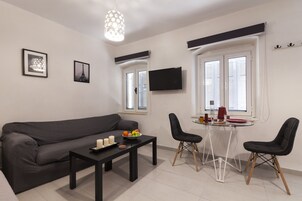 Uma sala de estar moderna com um sofá cinza, uma mesa de centro preta e uma mesa de jantar de vidro com duas cadeiras pretas. Há uma televisão de tela plana na parede e uma janela com persianas.
