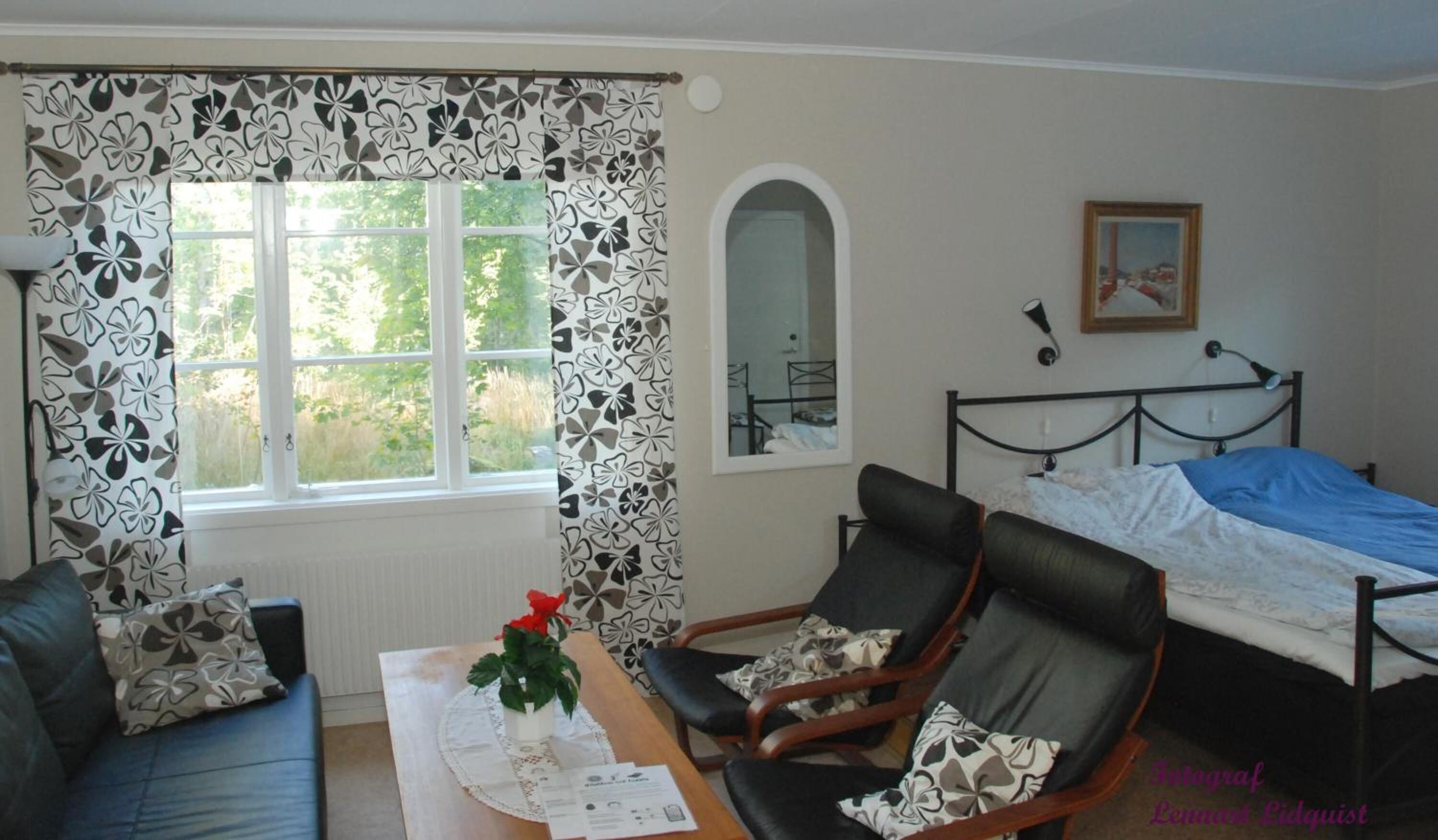Quarto com cama, escrivaninha com papéis, duas cadeiras, janela com cortinas estampadas e um quadro na parede.